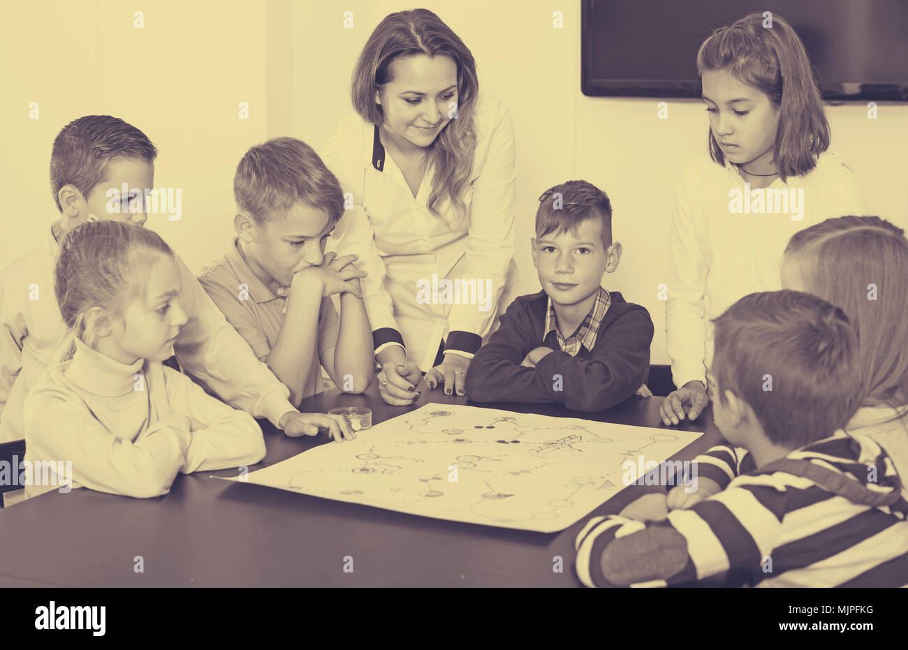 Female and happy kids sitting at table with board game in classroom ...