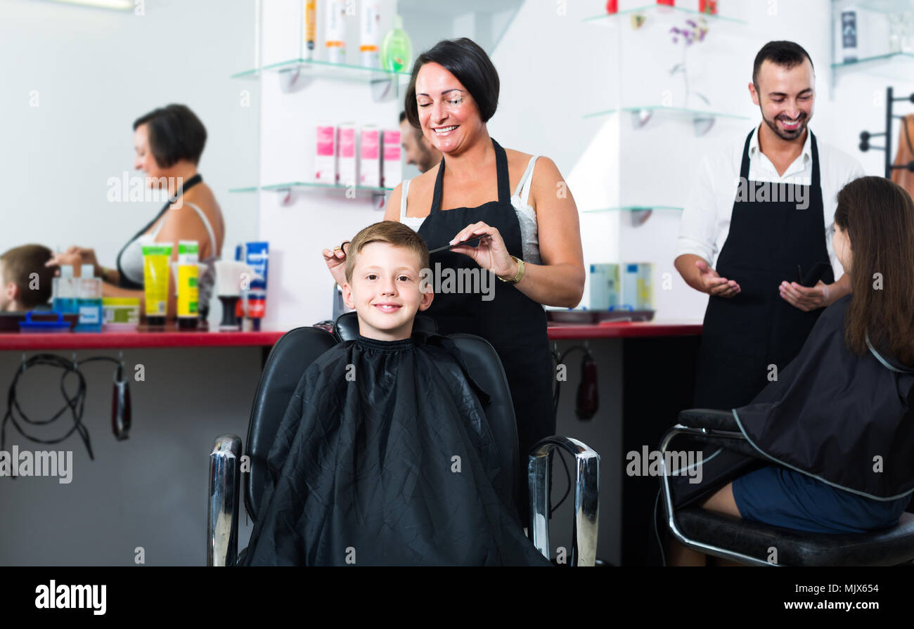 professional hairdresser in apron cutting hair of glad boy in crowded ...