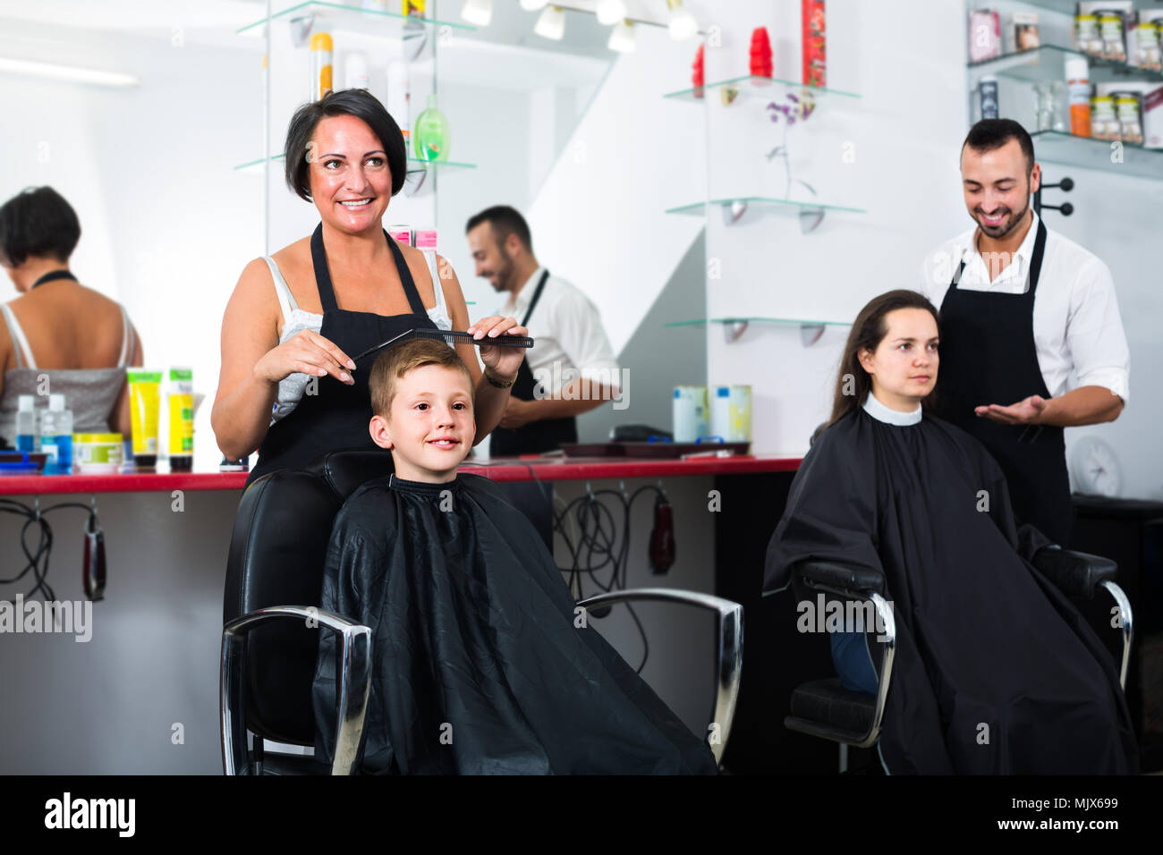cheerful woman hairdresser in apron cutting hair of glad customer in ...