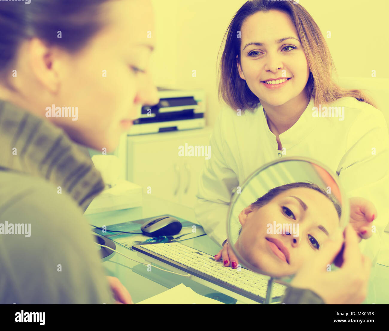 Young positive woman looking at mirror during consultation with doctor ...