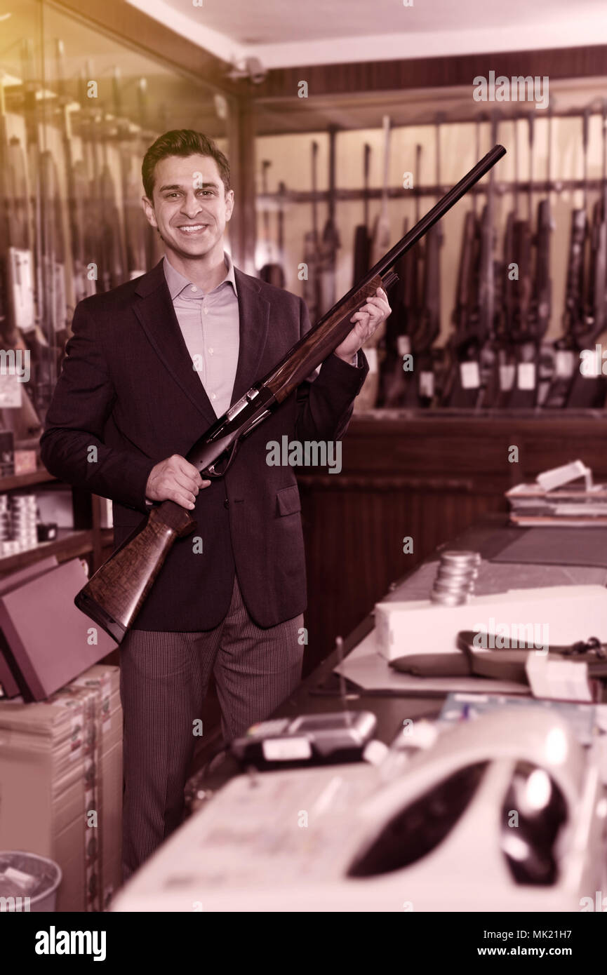 Portrait of positive male seller in hunting shop demonstrating shotgun ...