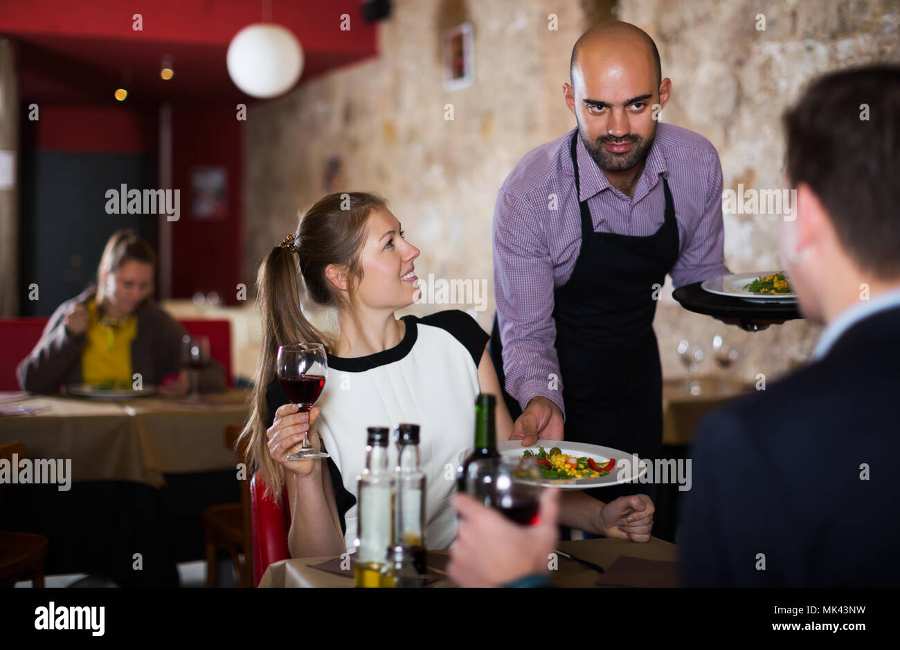 Polite waiter bringing ordered dishes to young couple at restaurant ...