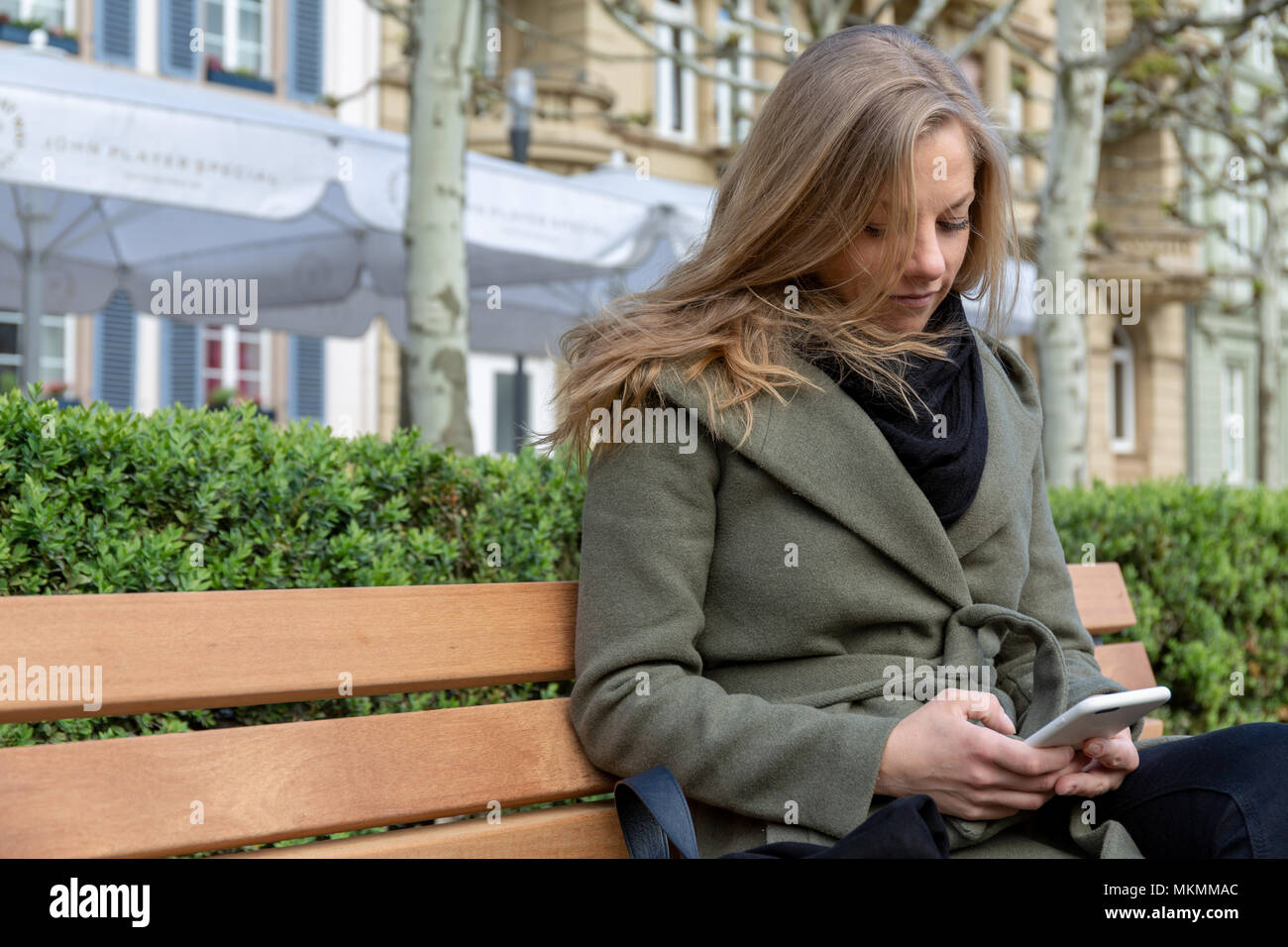 Young beautiful friendly woman sitting on a park bench wearing coat and ...