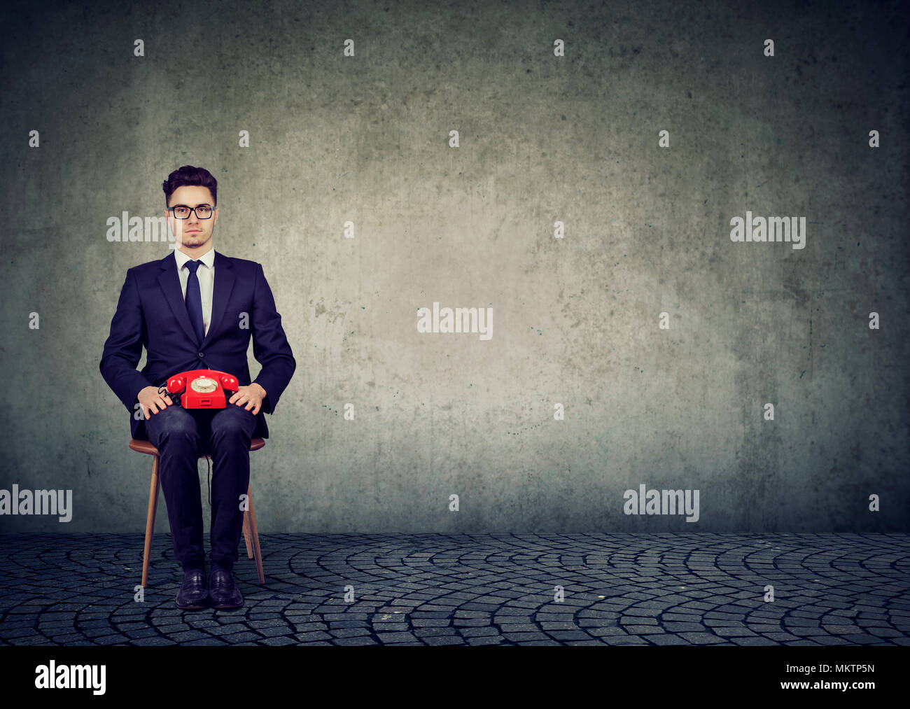 Young man in suit sitting with red telephone on chair against gray ...