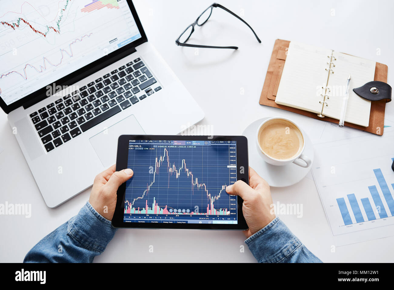 Young trader is sitting in home at table, working on tablet with graphs ...