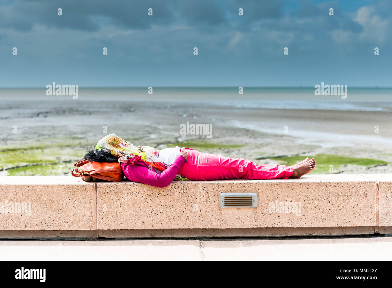 A sleepy tourist in colourful clothes takes a nap along the beach walk ...