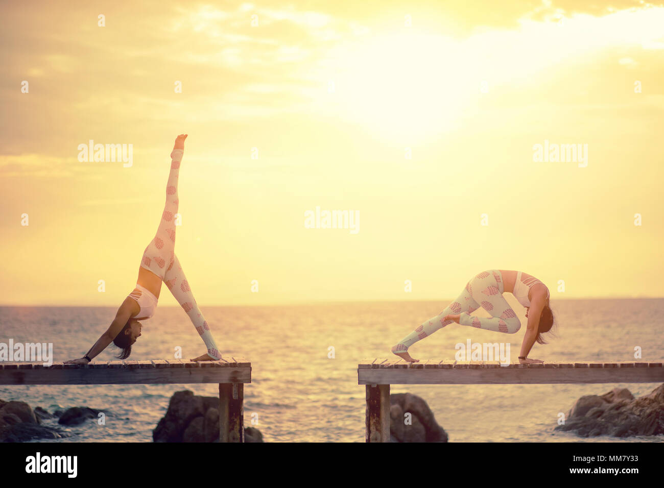 two woman playing yoga pose on wooden pier against sun rising over sea ...
