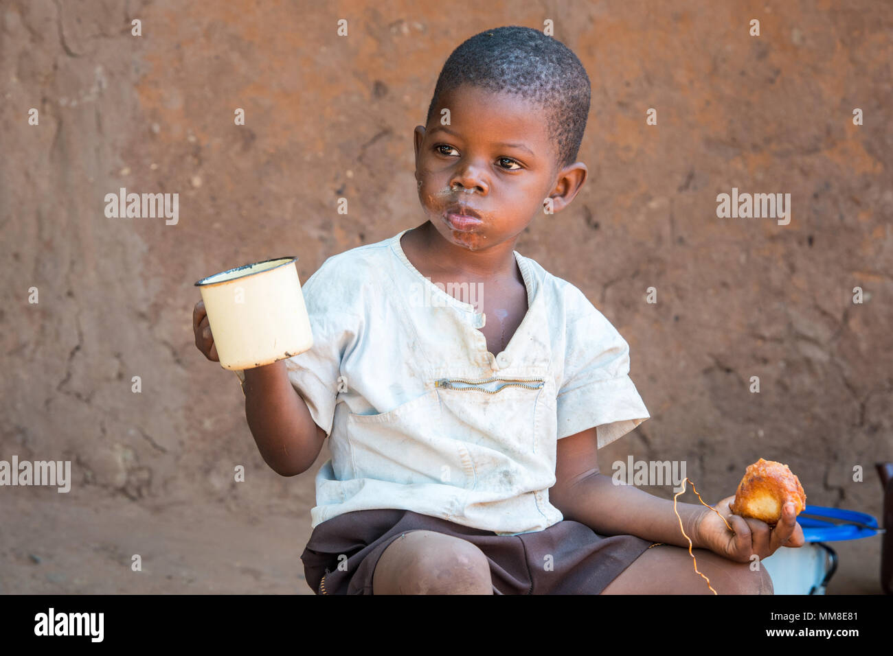 Zambian child snacks on fried pastry while holding a mug of water ...