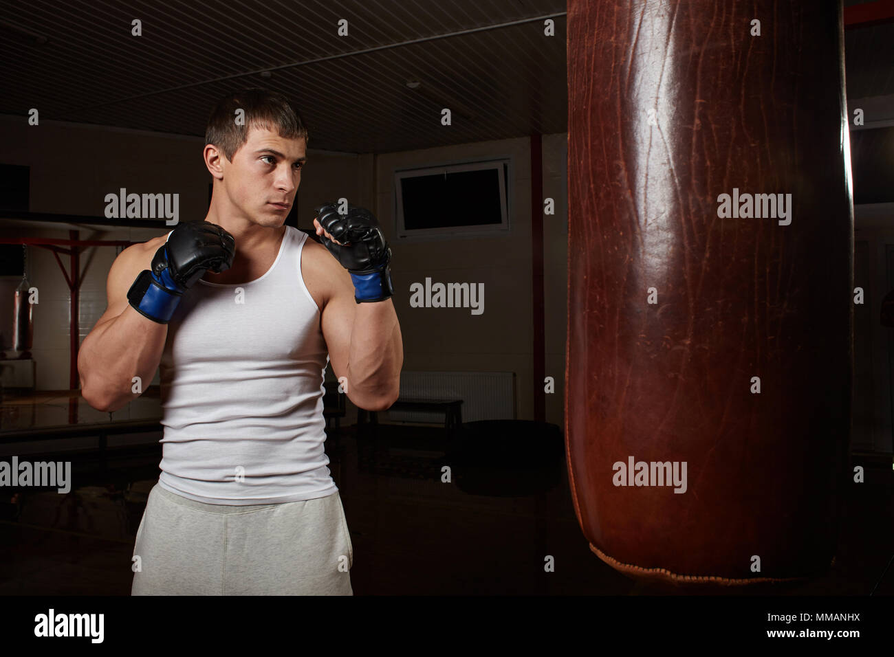 young muscular man boxer workout with punching bag in dark interior ...