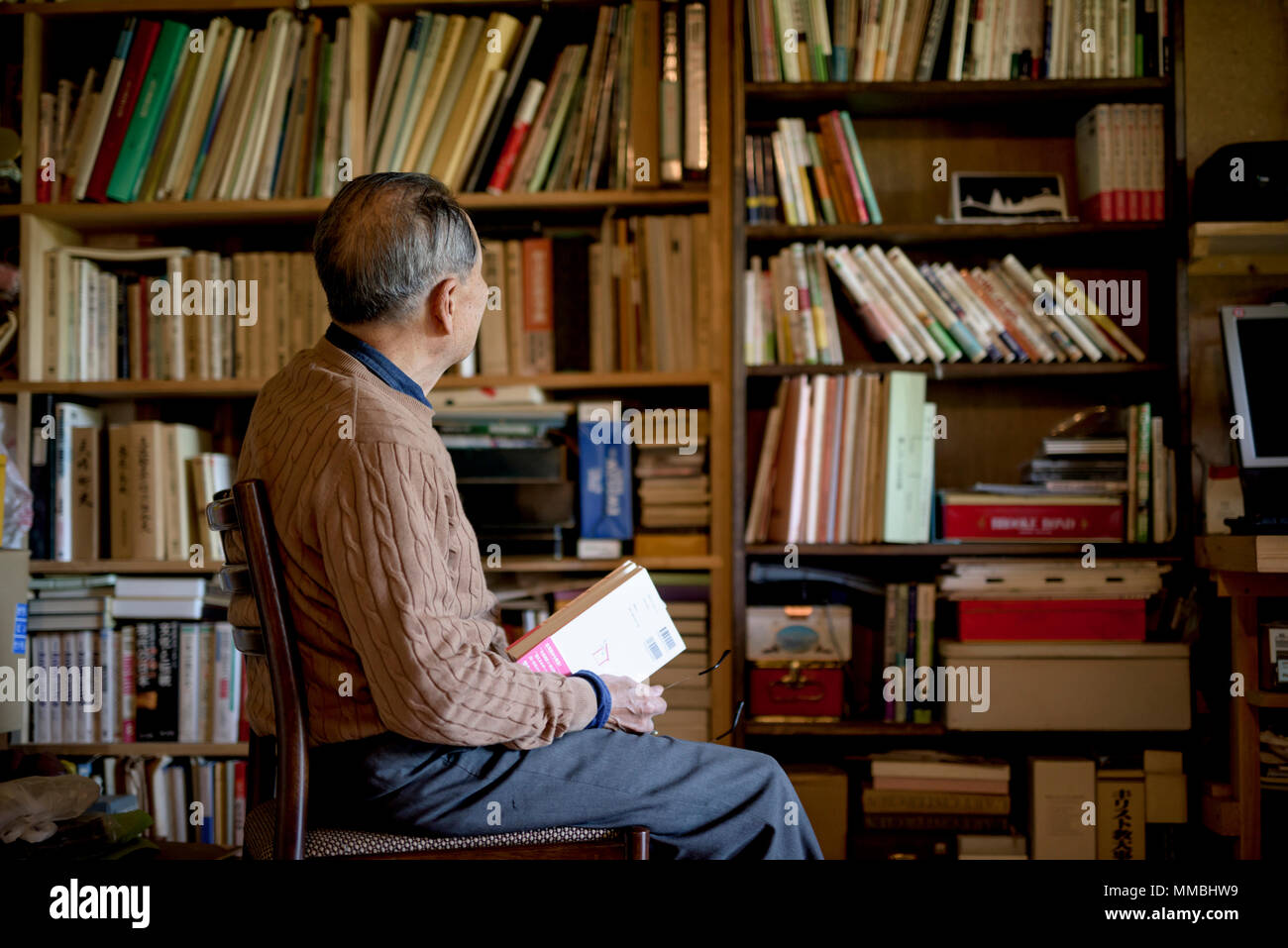Elderly man sitting on chair in front of bookcase, holding book Stock ...