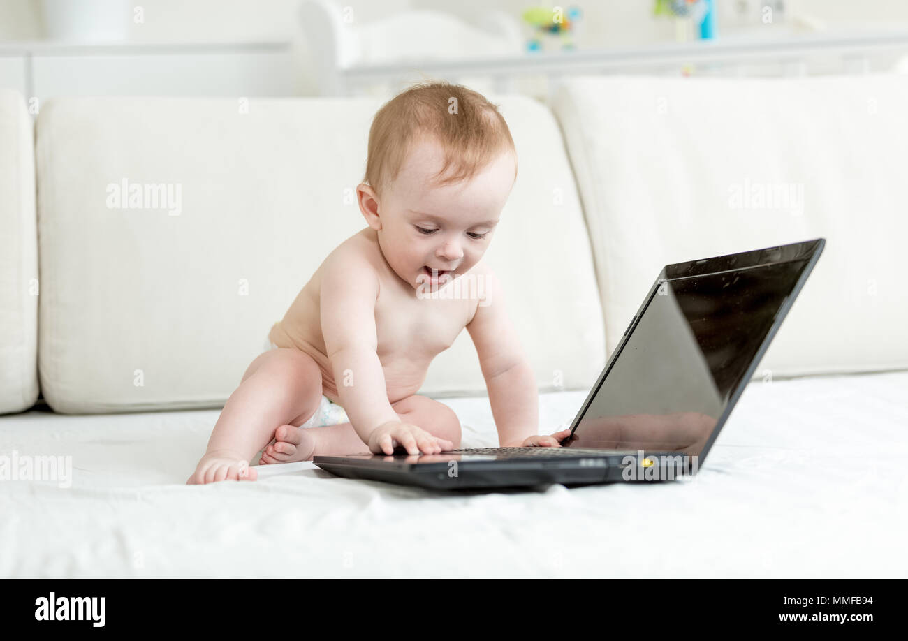 Happy smiling baby boy in diapers sitting on bed and using laptop ...