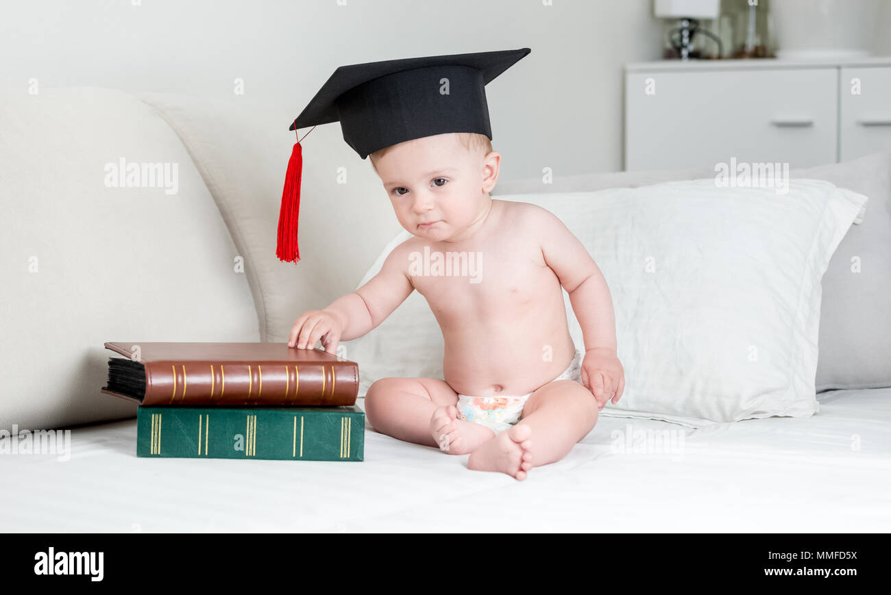 Cute baby boy in diapers and graduation cap sitting on bed with books ...