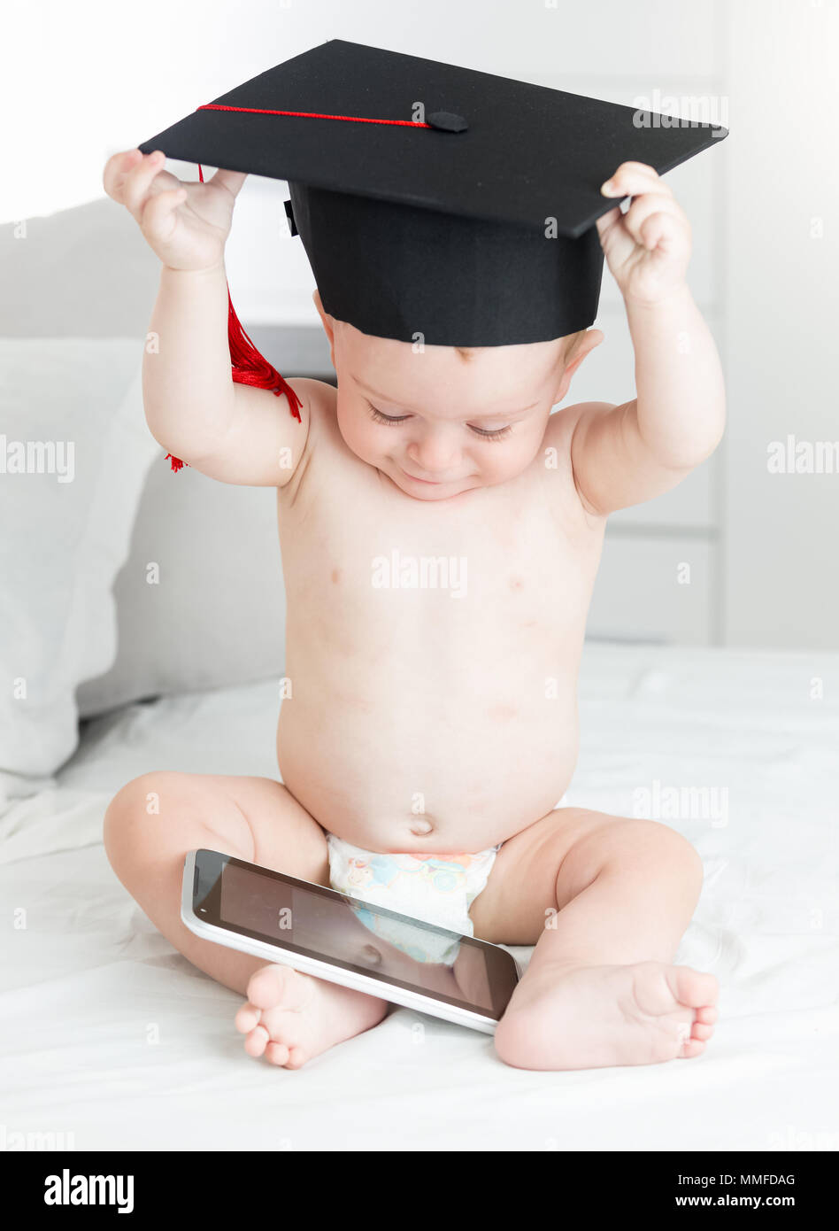 Portrait of baby boy in diapers and graduation cap sitting on bed and ...