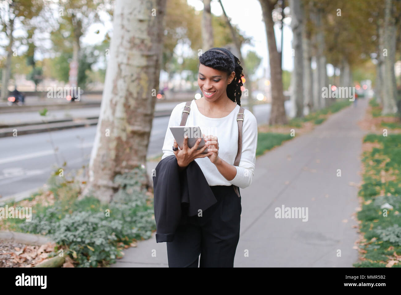 Black female student using tablet and walking on street with trees ...