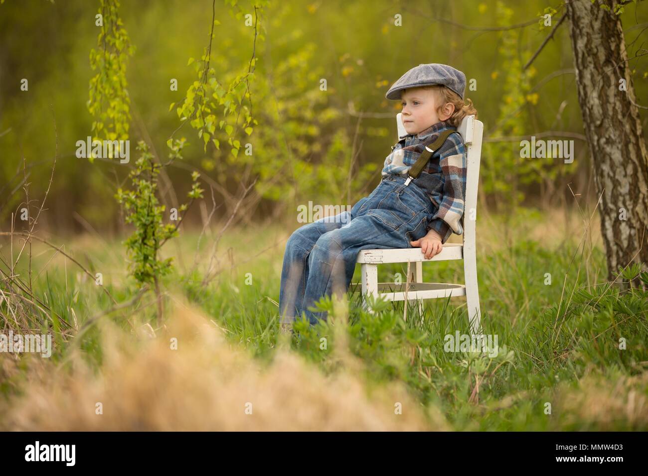 Young blonde boy relaxing on white old chair standing under birch tree ...
