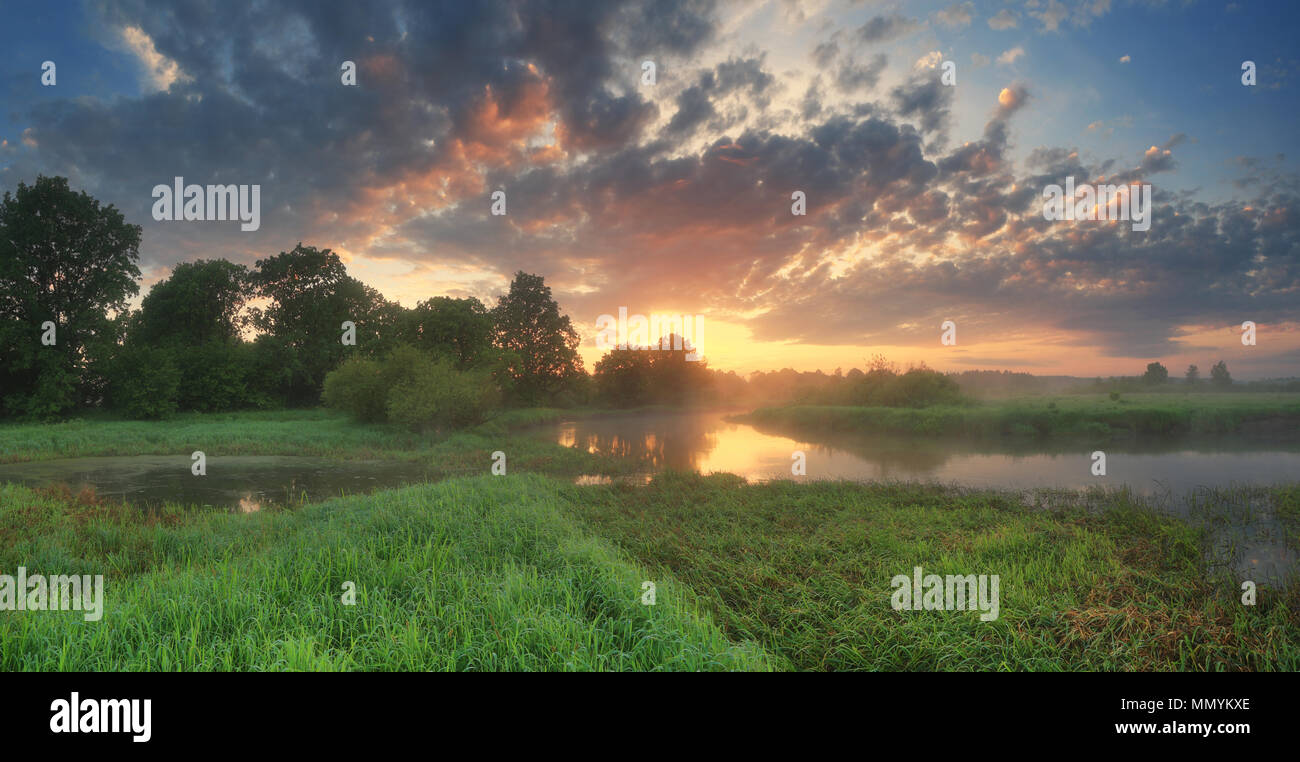 Early summer morning. Dramatic colorful sky over green field and lake ...