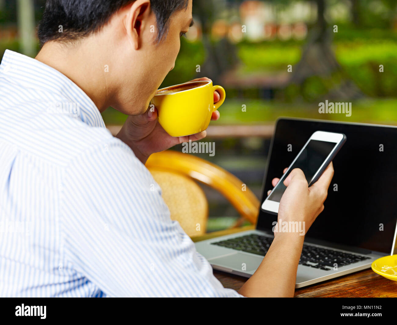 young asian man using mobile phone and laptop computer while drinking ...