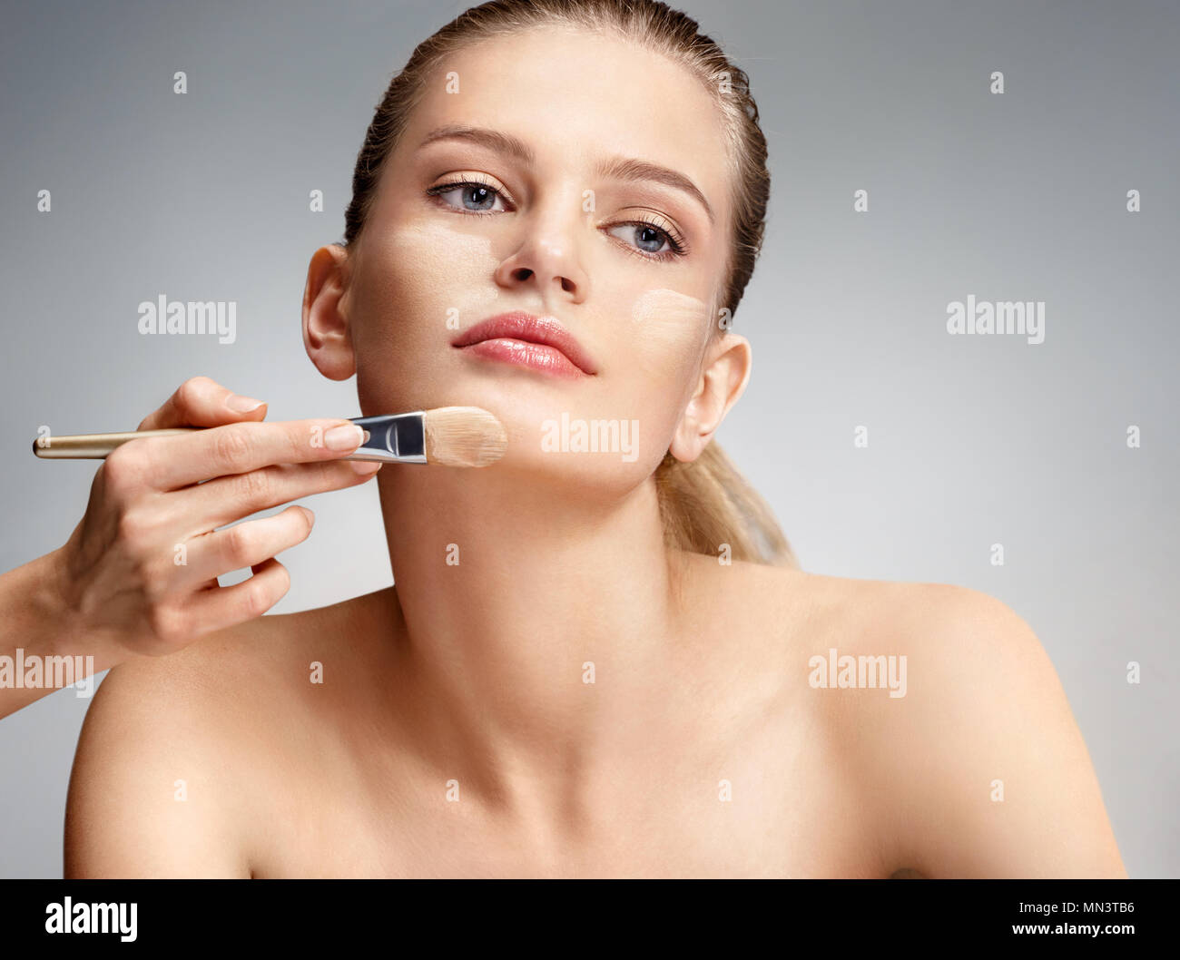 Beautiful woman applying liquid foundation on her face on grey ...