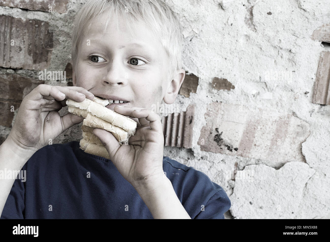 A hungry dirty boy greedily eats a crust of bread against the wall ...