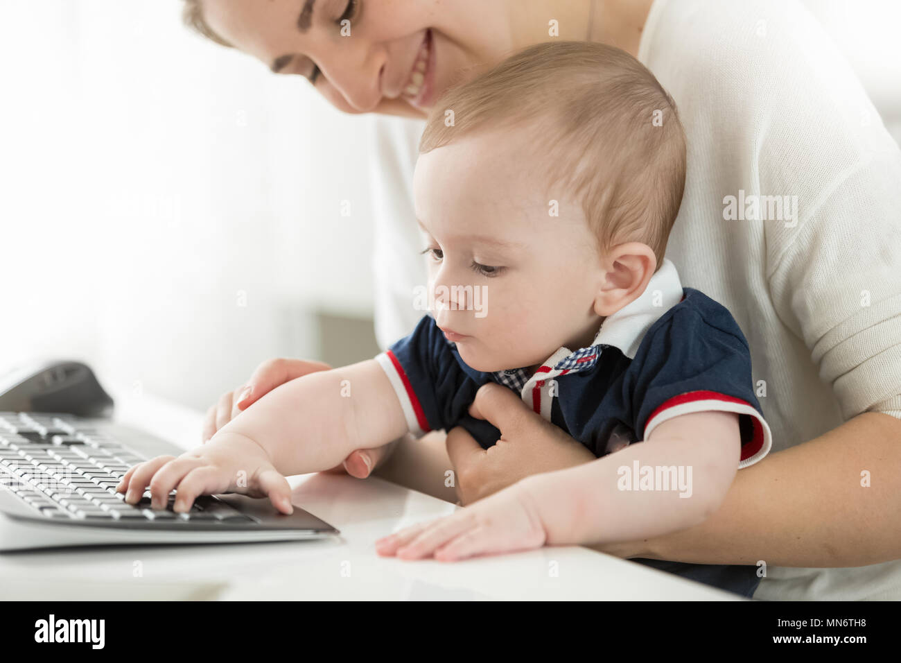 Cute baby boy sitting on mothers lap in office and typing on computer ...