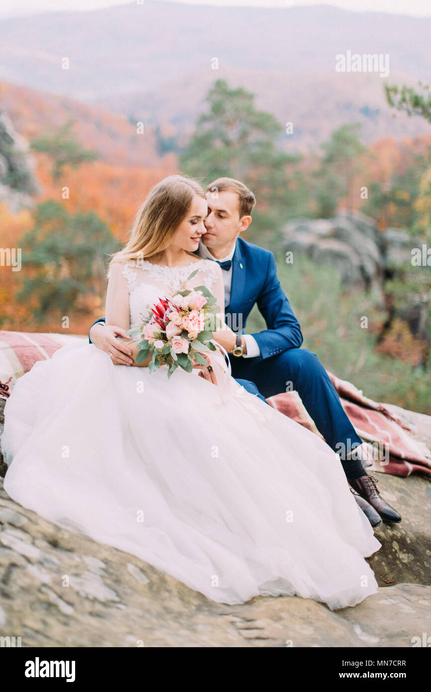 Romantic outdoor photo of the hugging newlywed couple sitting on the ...