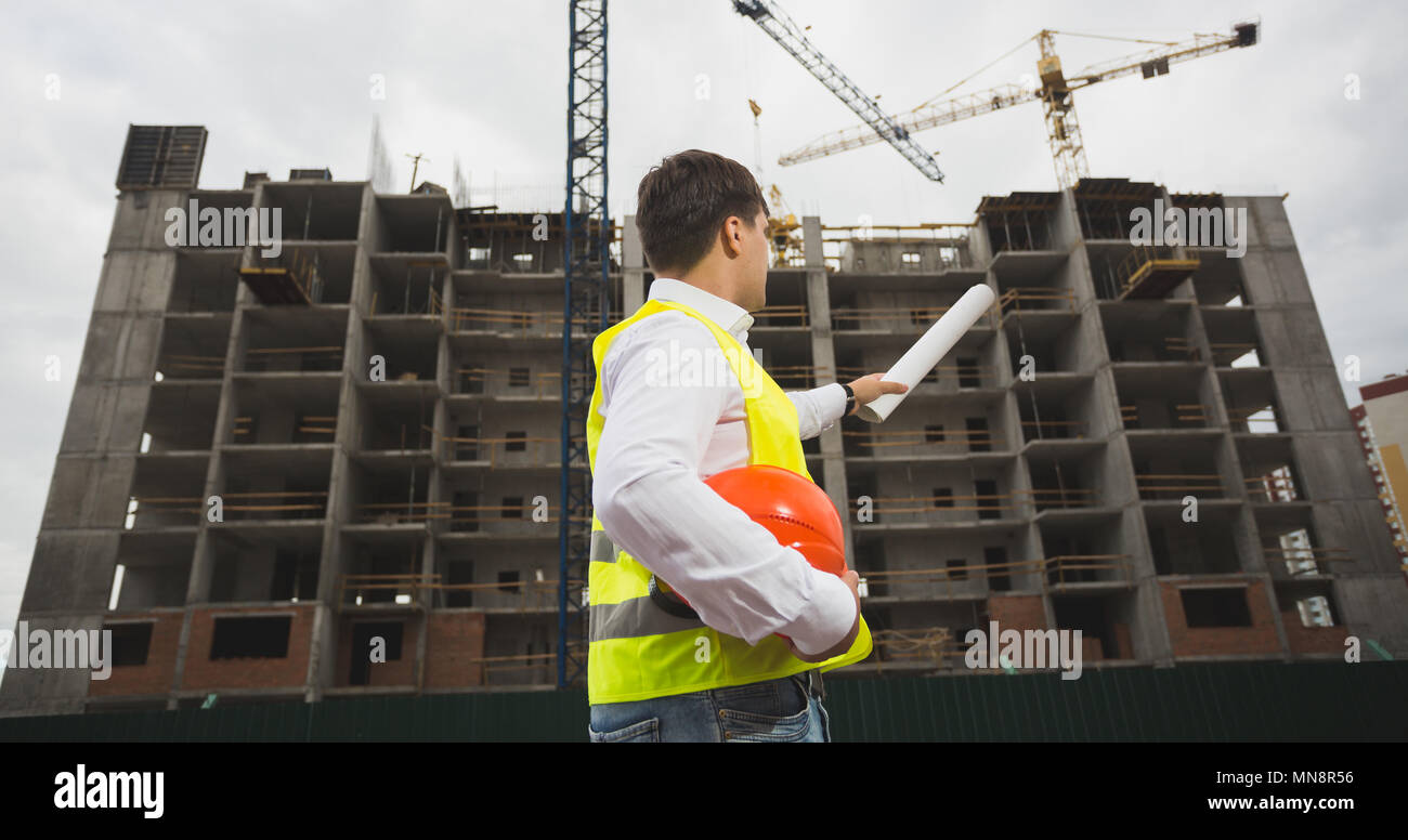 Rear view image of male architect in green safety vest showing building ...