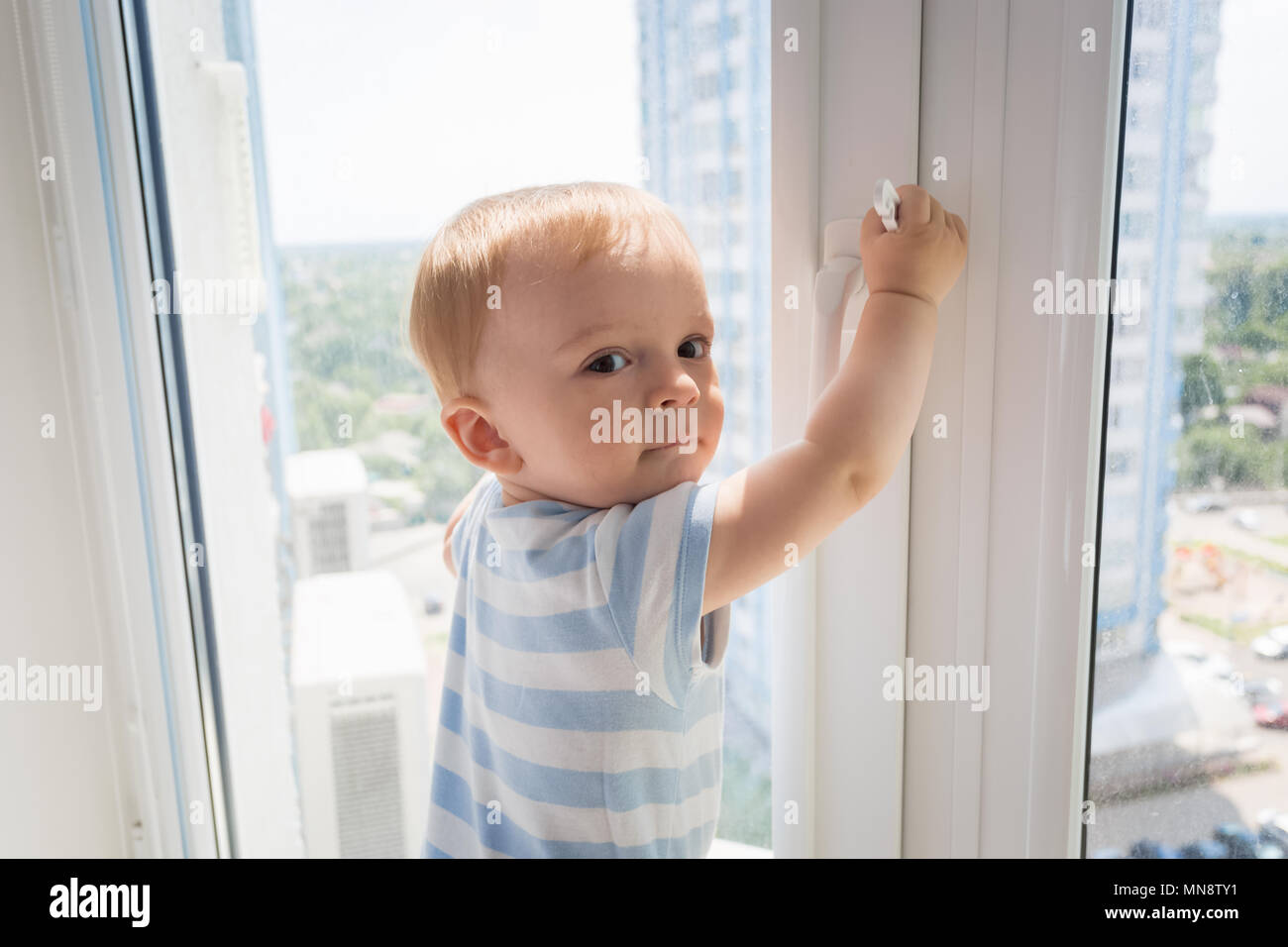 Portrait of little baby standing on window sill and trying to open the ...