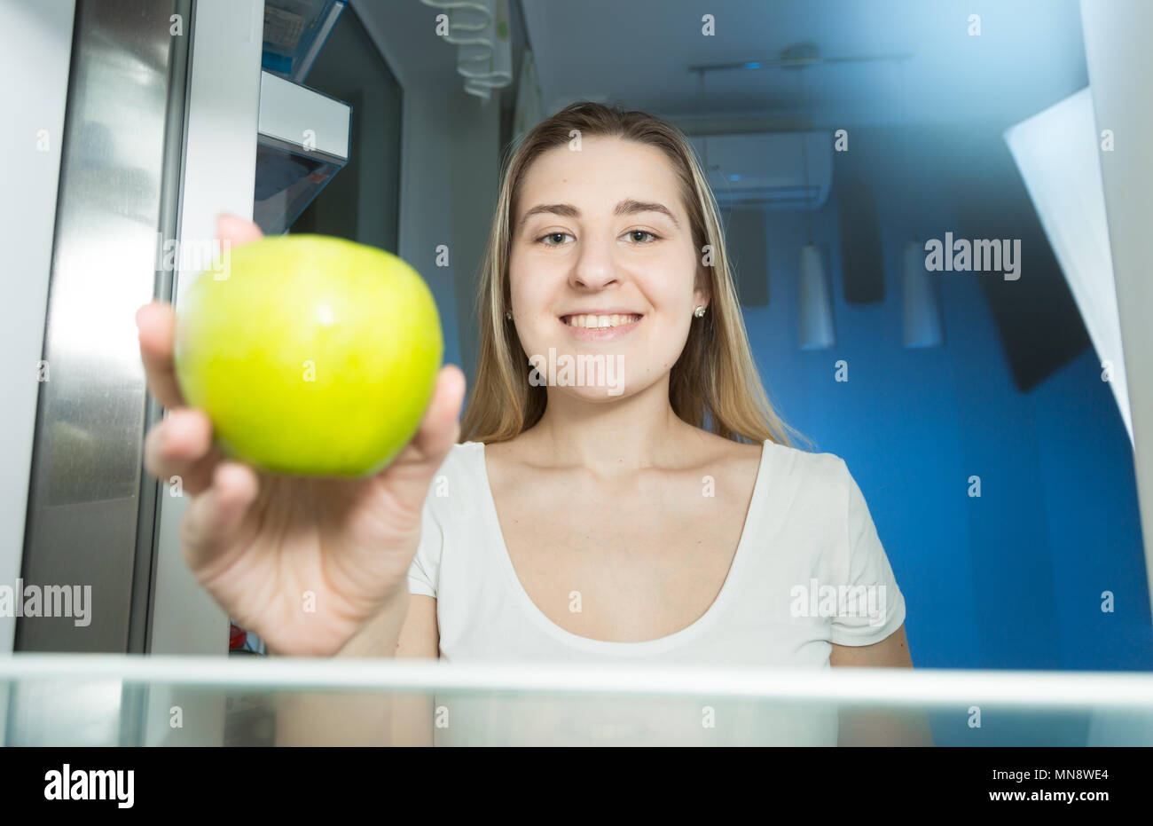 Young hungry woman taking fresh green apple from refrigarot shelf at ...