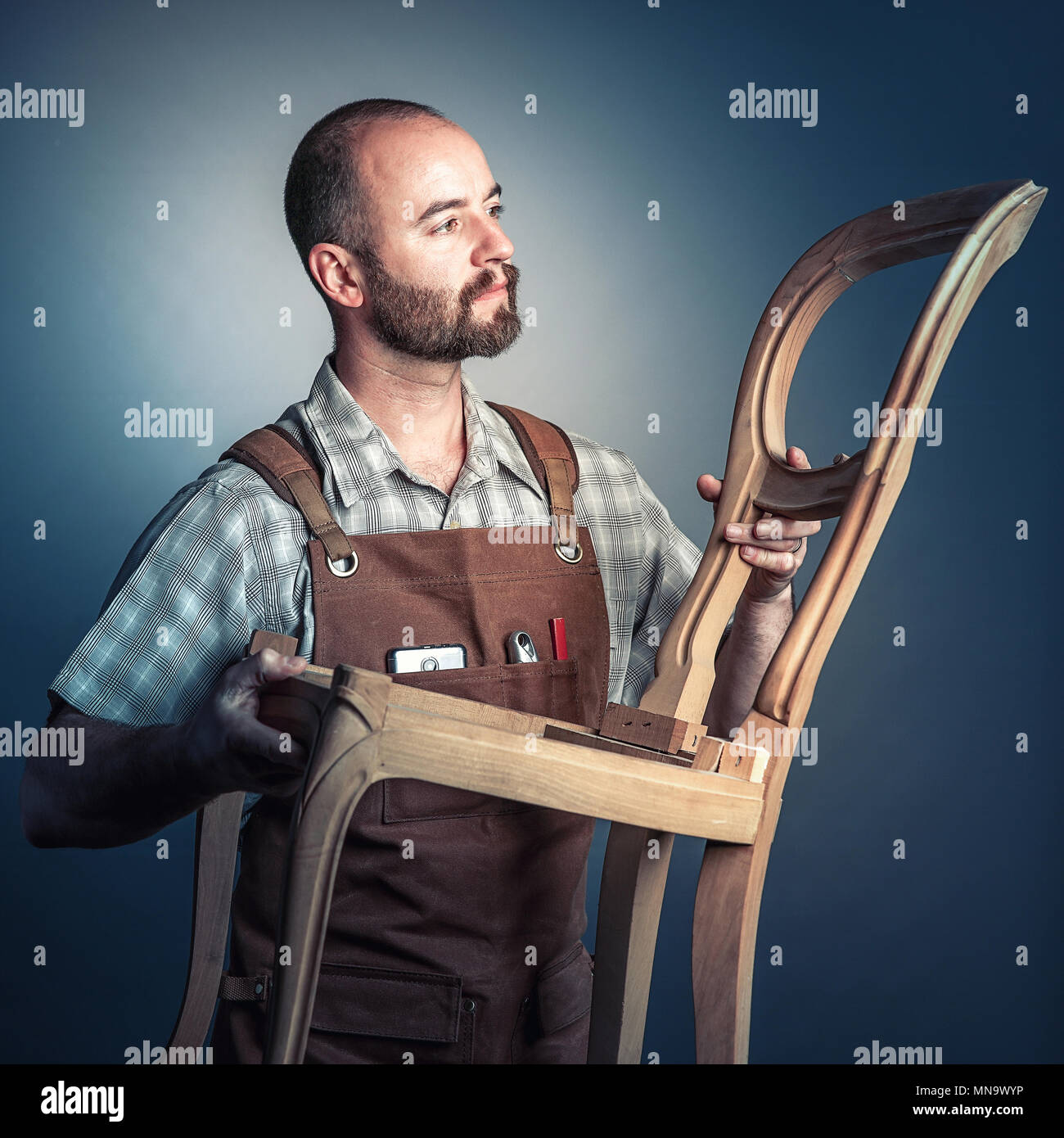 portrait of caucasian carpenter with apron and unfinished wood chair ...