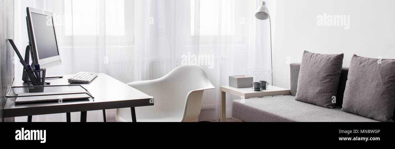 Desk with computer in spacious living room. Next to it grey comfortable ...