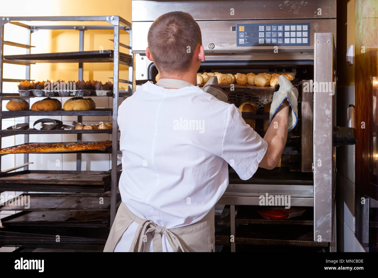 A male baker in white uniform and a beige apron bakes bread and takes ...