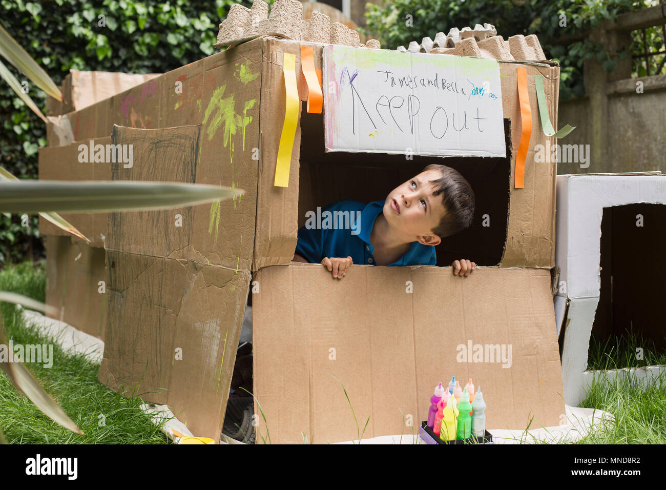 Cute boy playing in cardboard playhouse while looking up at back yard ...