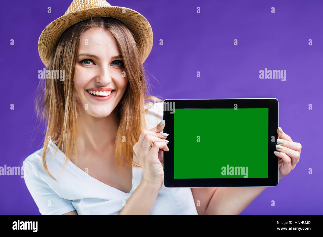 Smiling blonde girl dressed in white t-shirt and hat holds tablet ...