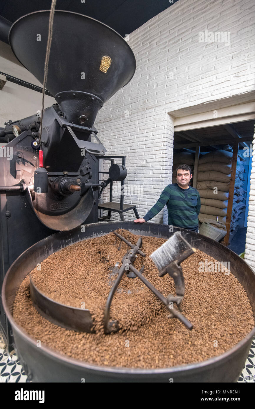 Adult male leans against coffee bean roaster as a mechanical arm slowly ...