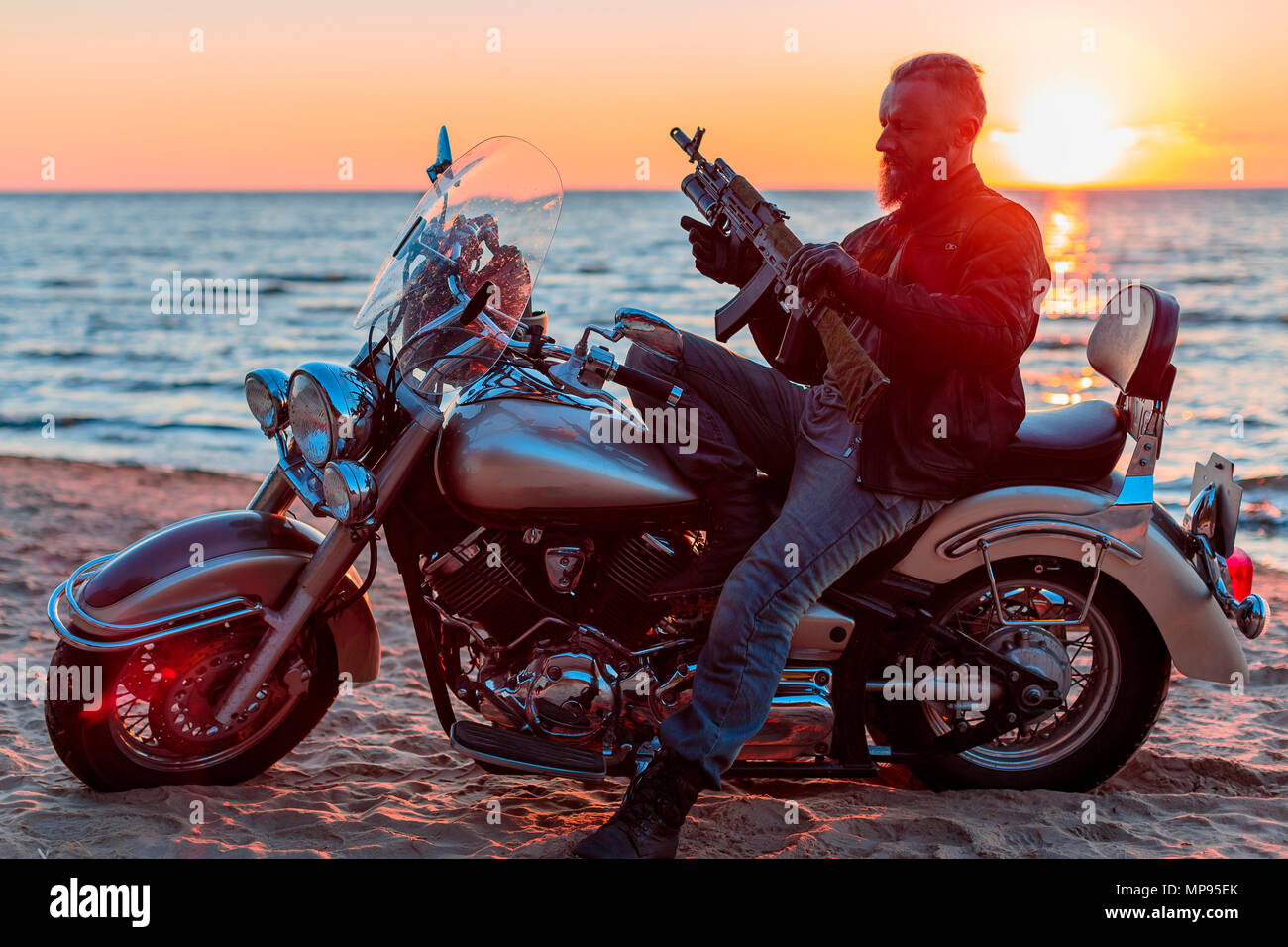 brutal man with a motorcycle on a sandy beach near the sea on a sunset ...