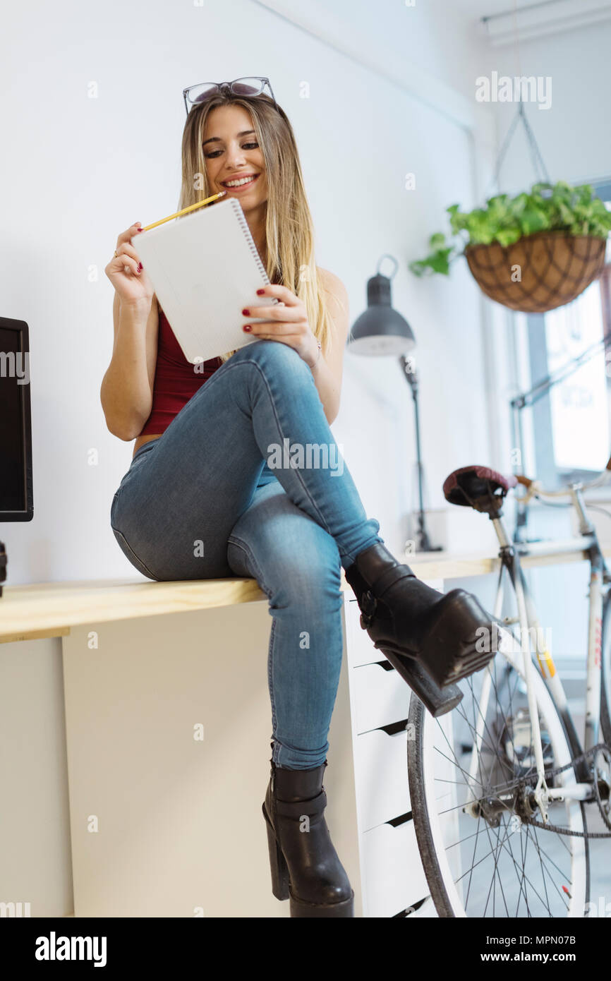 Smiling casual young woman sitting on desk in the office taking notes ...