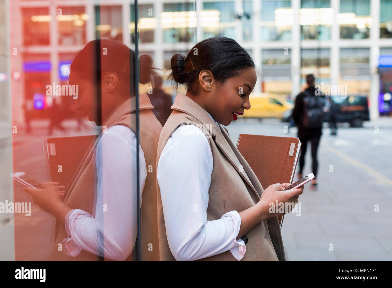 Smiling businesswoman leaning against glass pane using cell phone hi ...