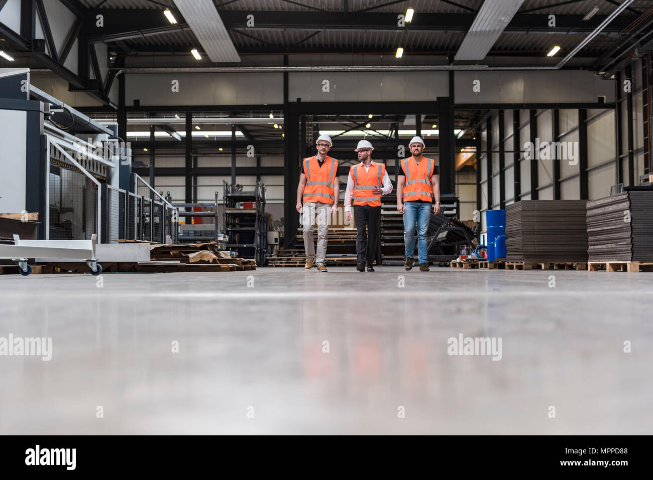 Three men wearing hard hats and safety vests walking on factory shop ...