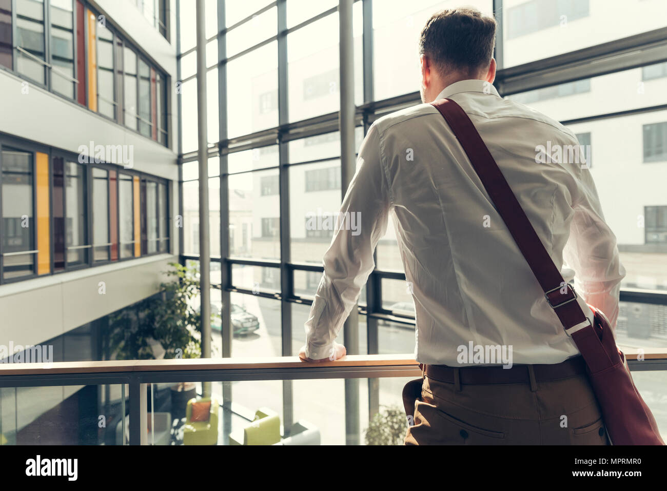 Rear view of businessman standing in office building leaning on railing ...
