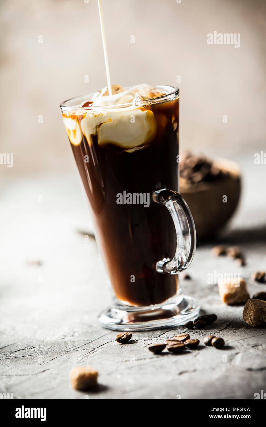 Ice coffee in a tall glass with cream poured over and coffee beans on a grey stone background ...