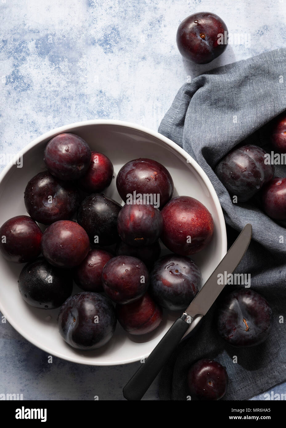 A bowl of ripe red plums and a knife with scattered plums on a blue ...
