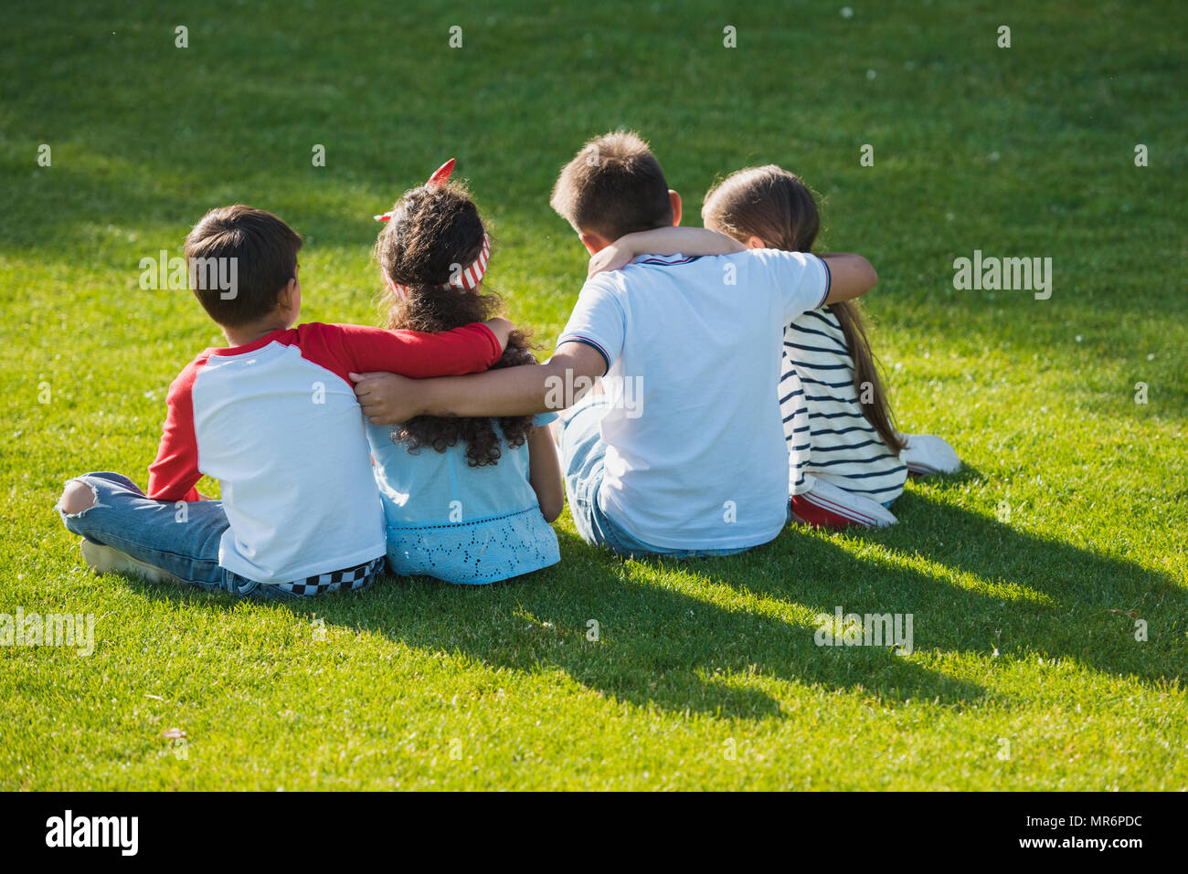 Back view of cute kids sitting embracing on green grass in park Stock ...