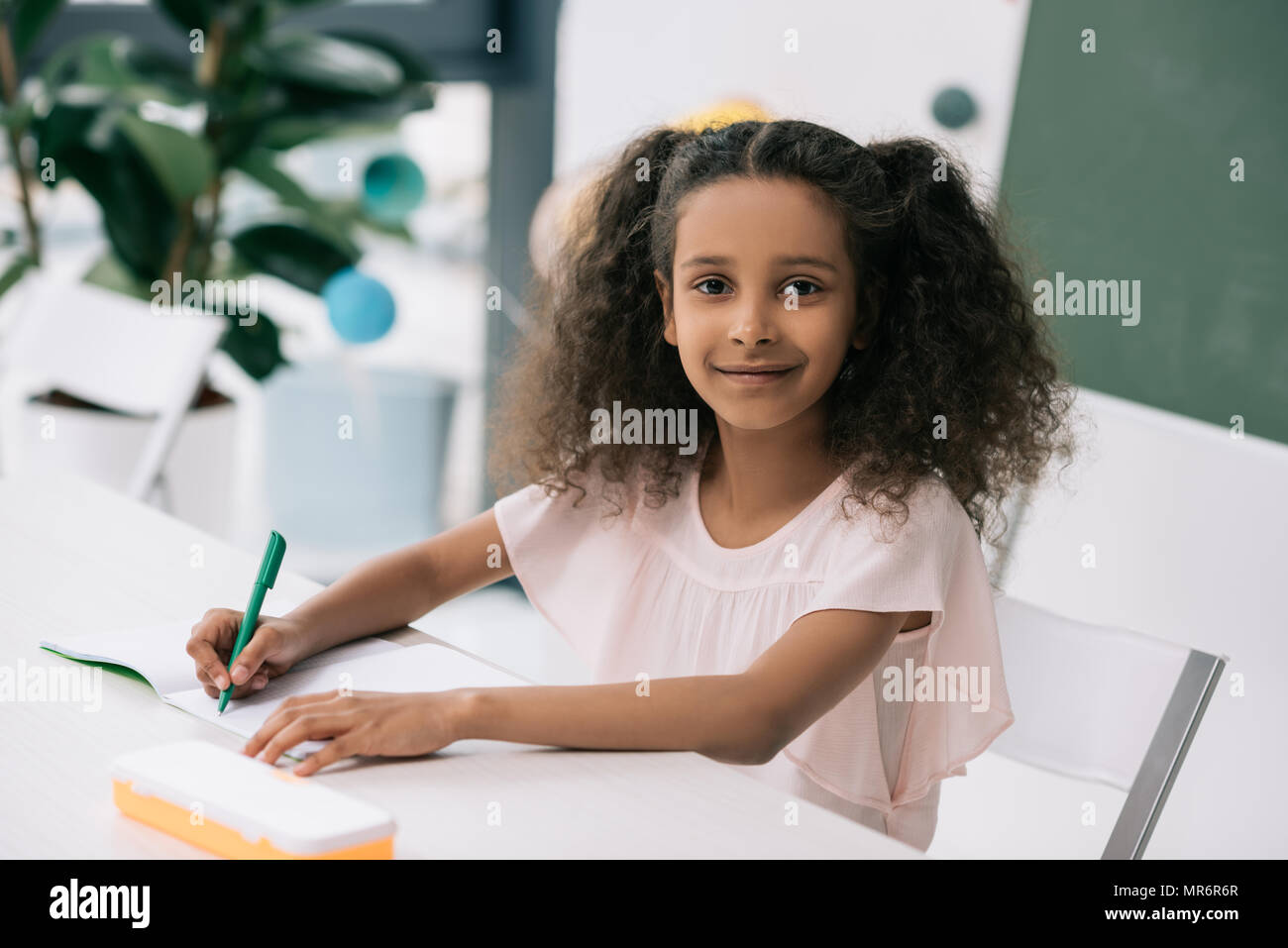 Cute african american pupil writing in exercise book and smiling at ...