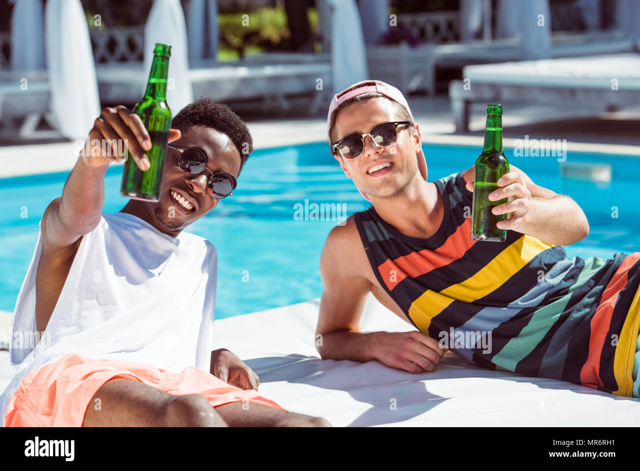 Two happy multiethnic men sitting with beer near swimming pool at ...