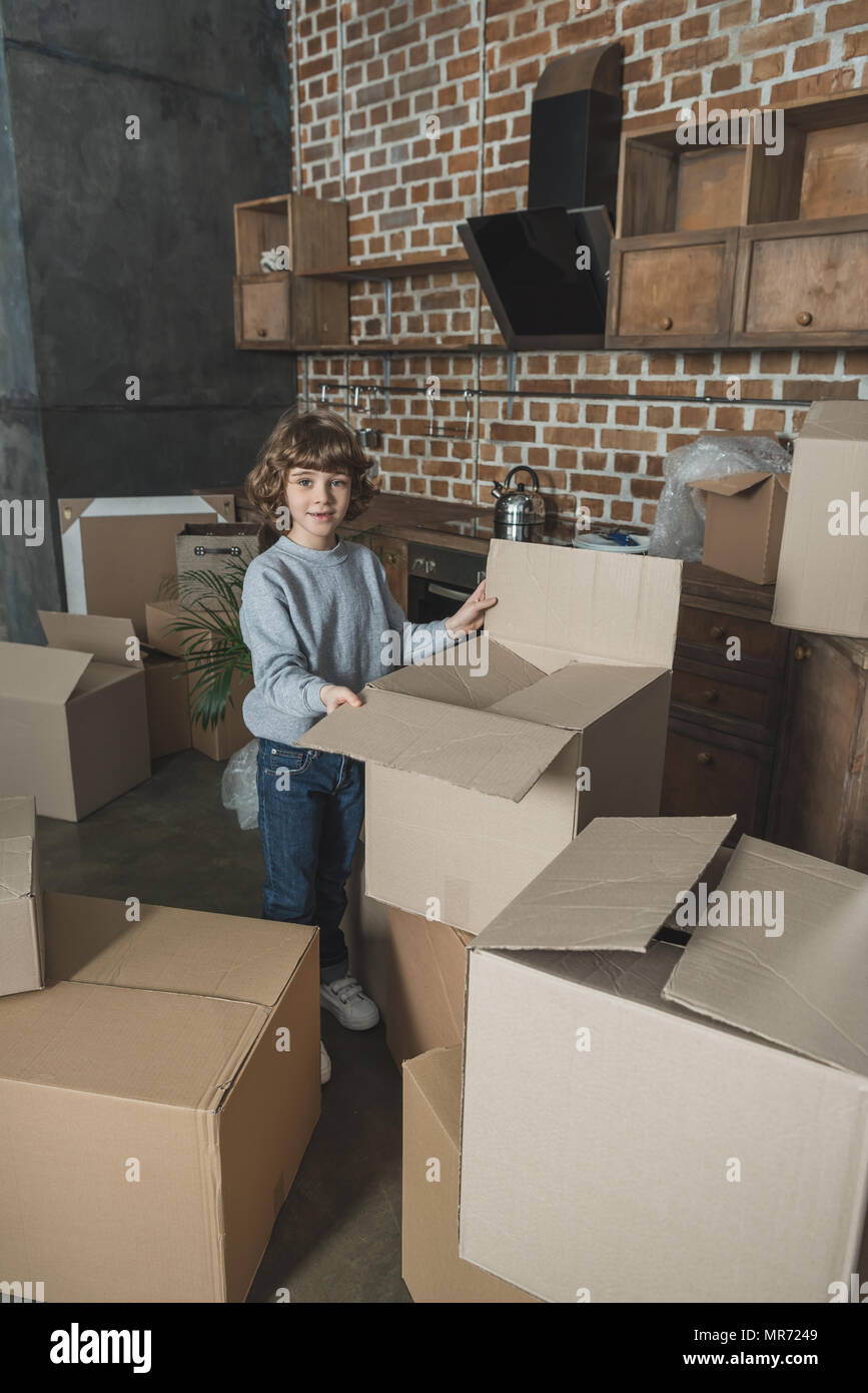 cute little boy smiling at camera while packing boxes during relocation ...
