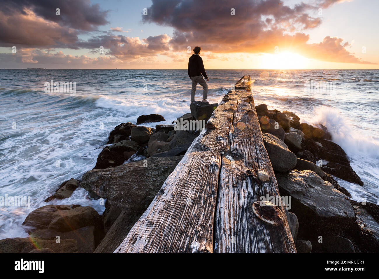A man stands on a jetty looking out towards the setting sun and wild ...