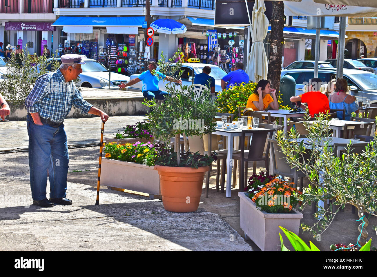 An old local Greek man with walking stick looks at people relaxing in a ...