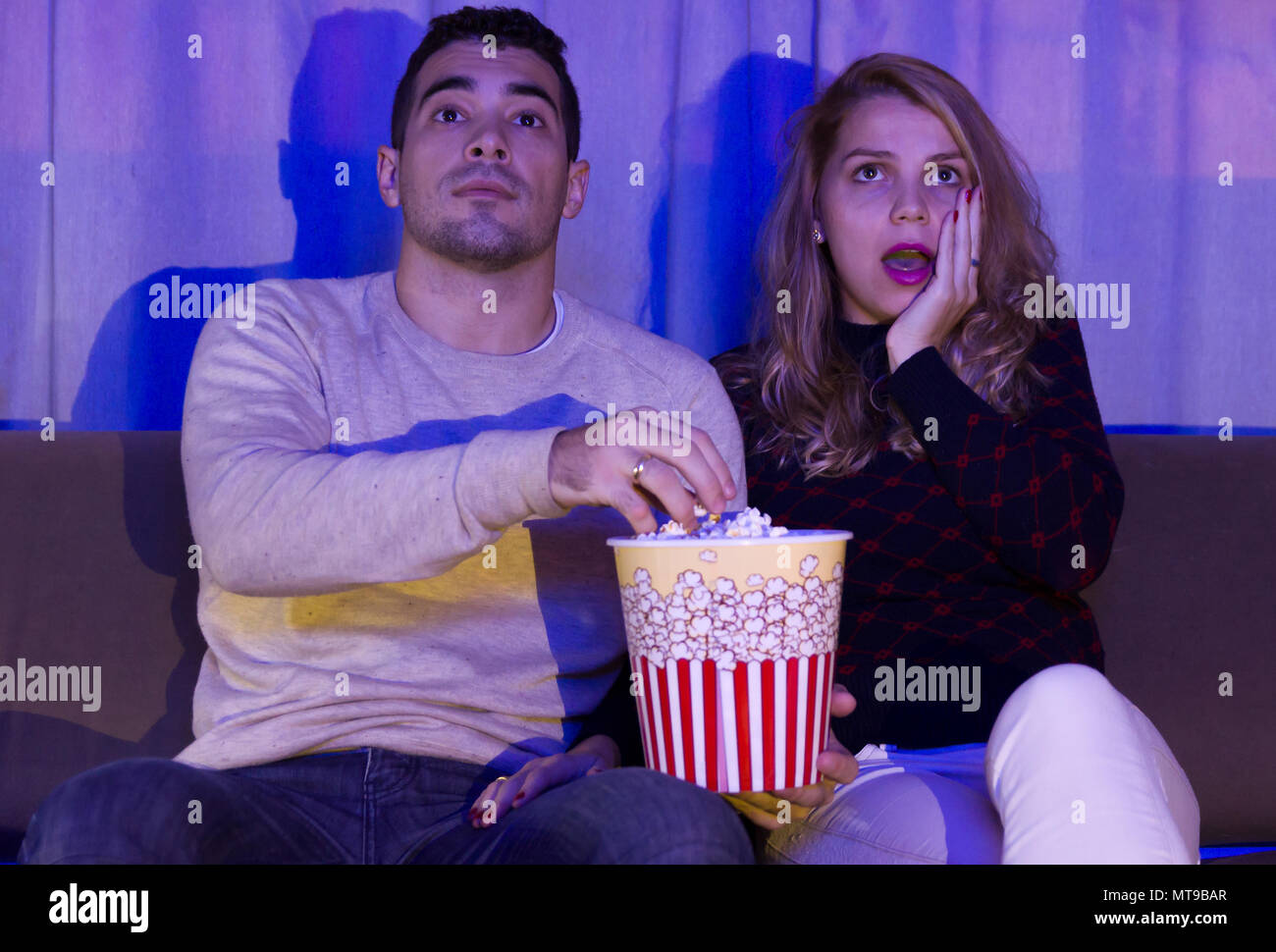Young Couple Watching Thriller Movie In Dark Room With Popcorn On Sofa