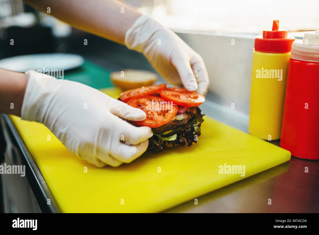 Male cook puts tomatoes in to the burger, fast food cooking. Hamburger ...
