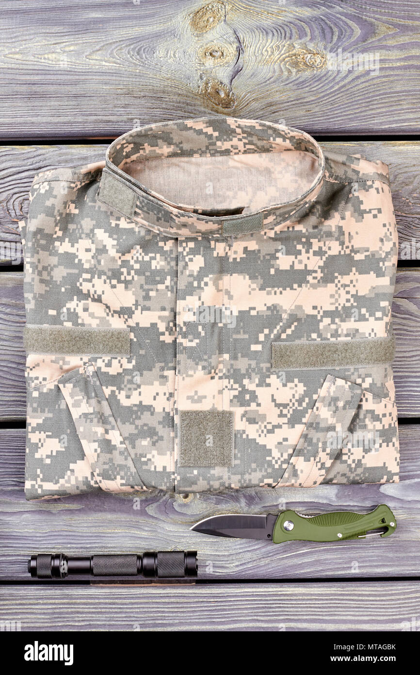 Folded camouflage jacket, flashlight, and knife. Wooden desk surface ...